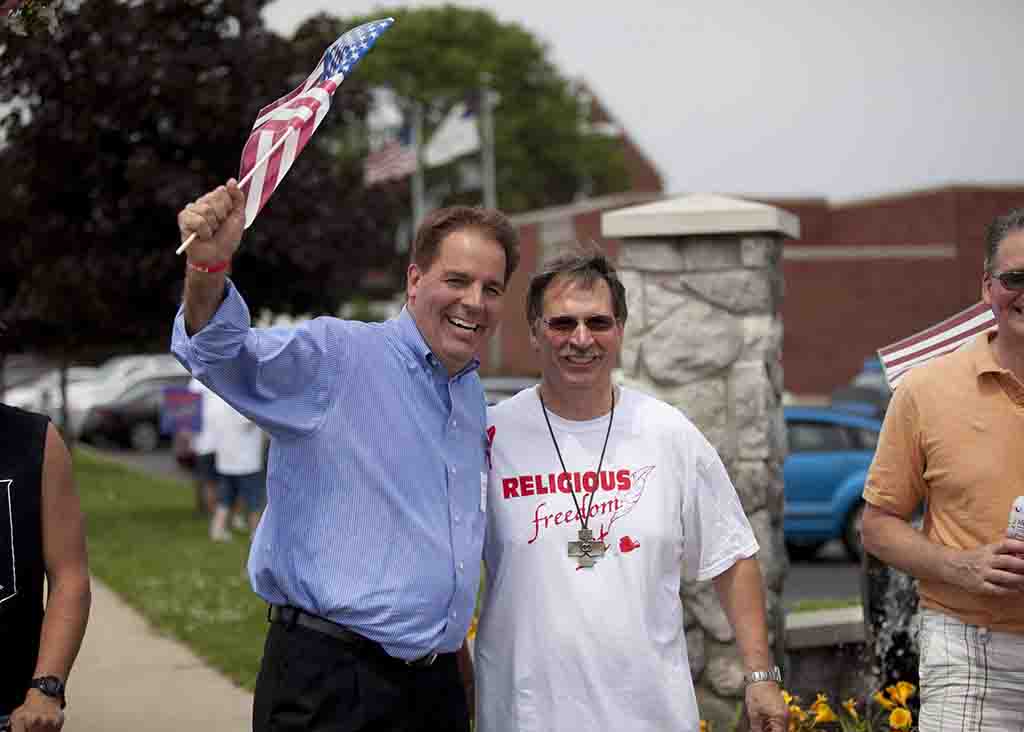 Rick clutches an American flag as he poses with a man wearing a shirt ...